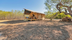 Uma estrutura rústica em forma de tenda, com uma escada que leva a uma plataforma, cercada por grama seca e árvores esparsas.