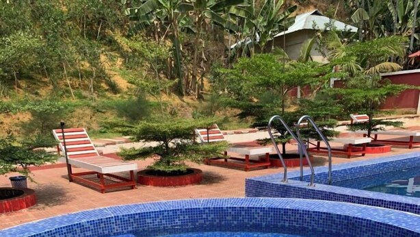 Uma piscina circular com azulejos de mosaico azul, rodeada por um espaço de deck vermelho e branco com espreguiçadeiras e uma pequena árvore.