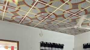 Um quarto com piso xadrez, cama com roupa de cama branca, duas cortinas listradas em preto e branco, uma cadeira de vime e um teto com padrão geométrico.