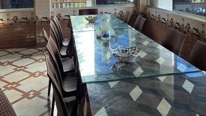 Uma sala de jantar espaçosa com mesa de vidro e cadeiras de vime, janelas amplas com vista para o verde e um ventilador de teto.