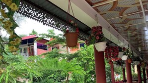 Uma área externa coberta com piso xadrez, balaústres vermelhos e brancos, vasos de flores pendurados e vegetação exuberante ao fundo.