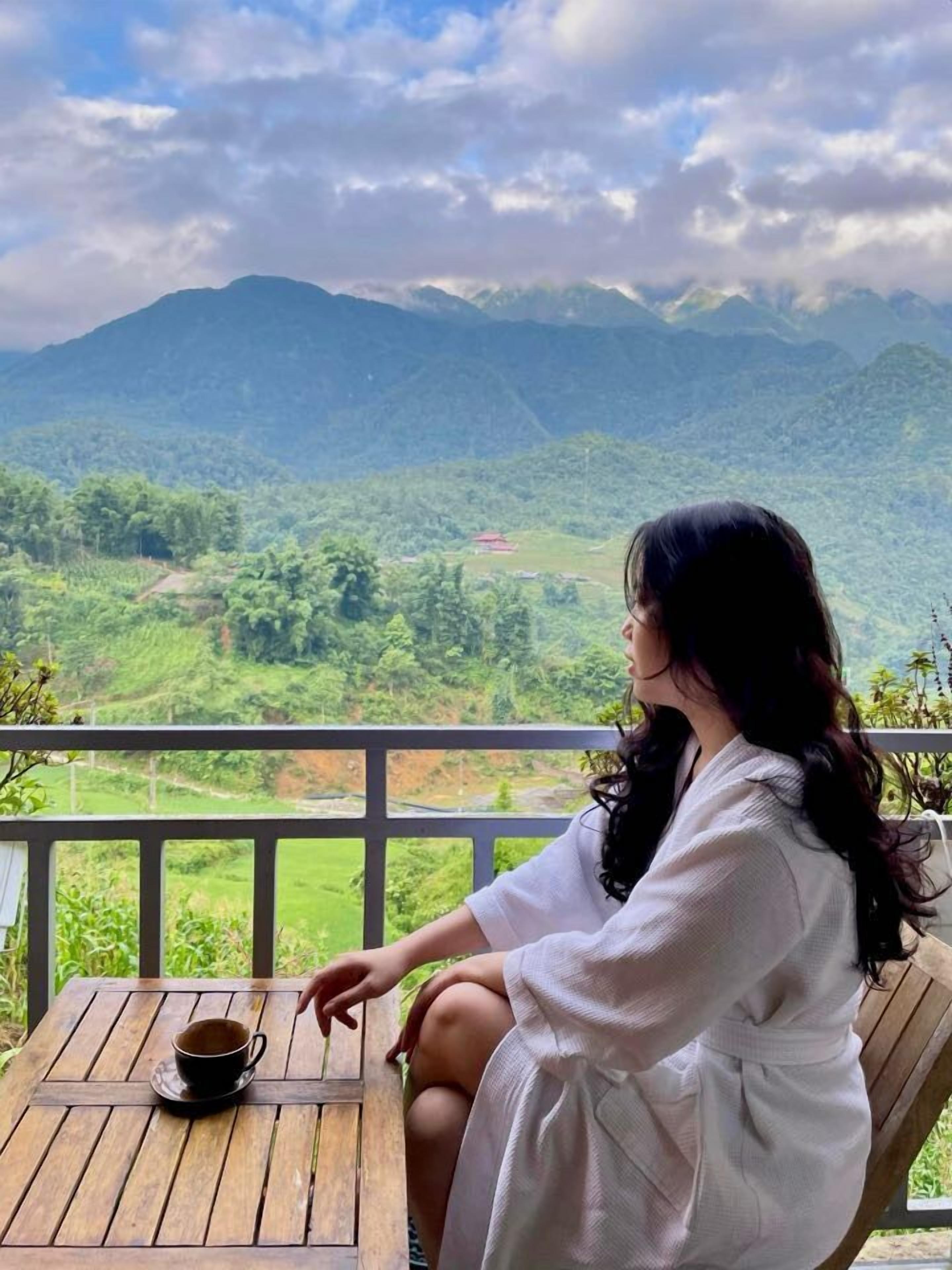 Uma pessoa sentada à mesa de madeira com uma xícara, apreciando a paisagem montanhosa.