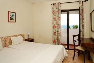 Quarto com cama, escrivaninha, cadeira, mesa de cabeceira e varanda com cortinas.