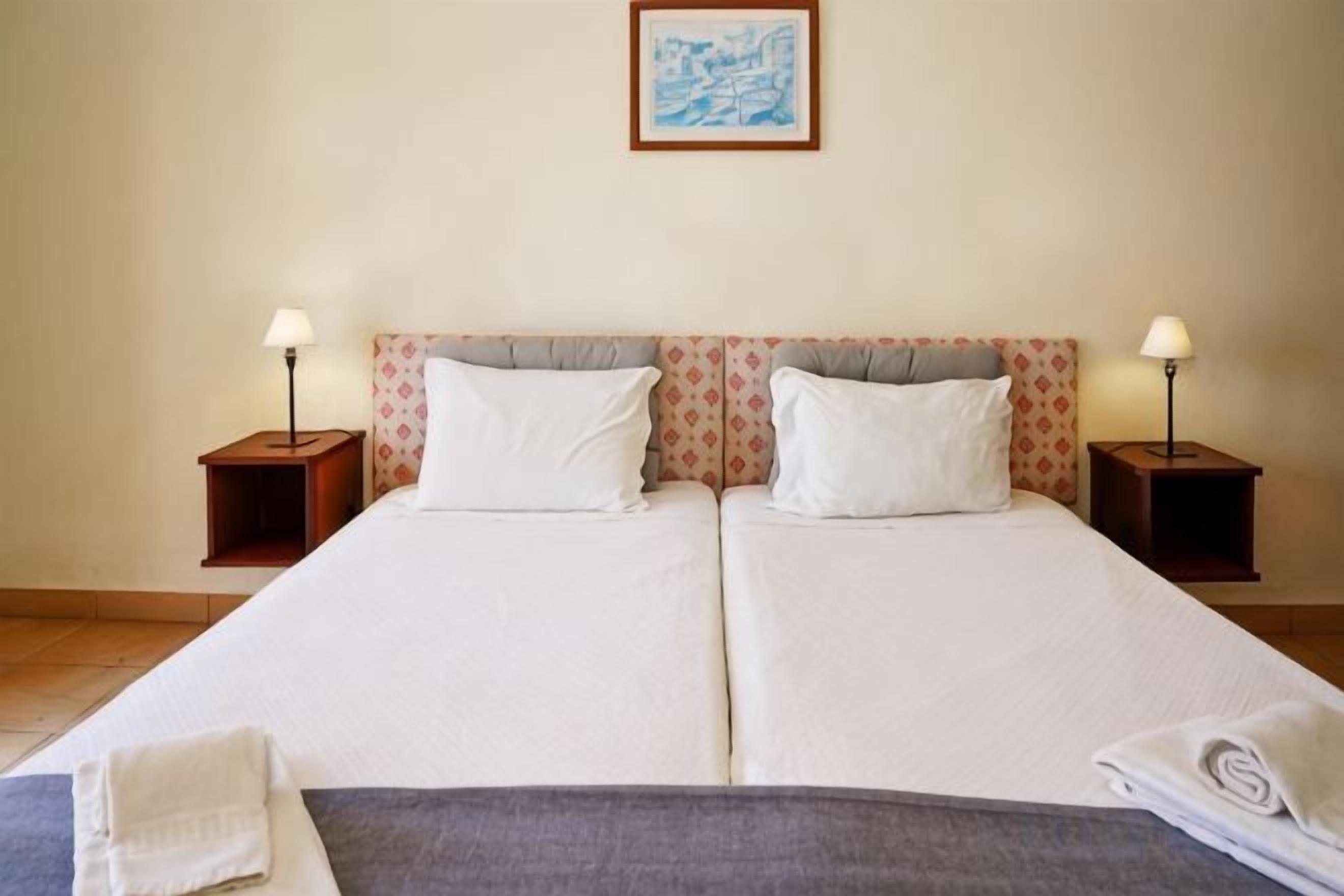 Cama de casal com duas almofadas, mesa de cabeceira com abajur e quadro na parede.