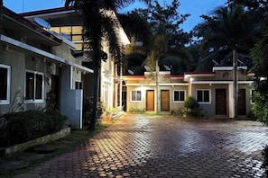 Um pátio pavimentado com um edifício à esquerda, uma área coberta com portas à direita e uma palmeira no centro.
