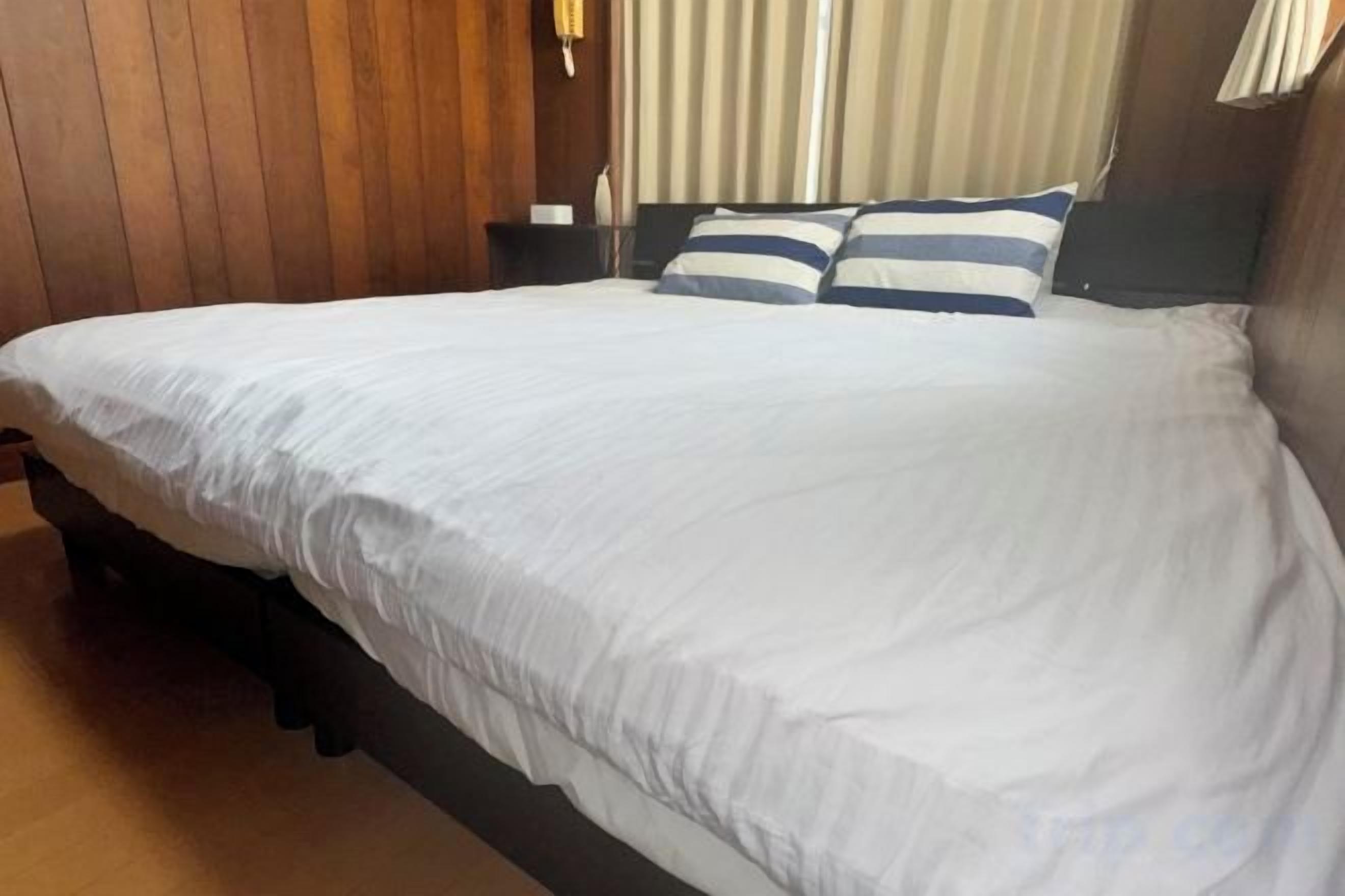 Uma cama com colcha branca e duas almofadas, cabeceira de madeira e mesa de cabeceira com abajur.