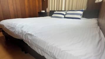 Uma cama com colcha branca e duas almofadas, cabeceira de madeira e mesa de cabeceira com abajur.