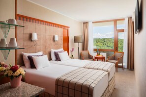 Quarto de hotel com duas camas, área de estar com mesa e cadeiras, televisão na parede e janela com vista para vegetação.