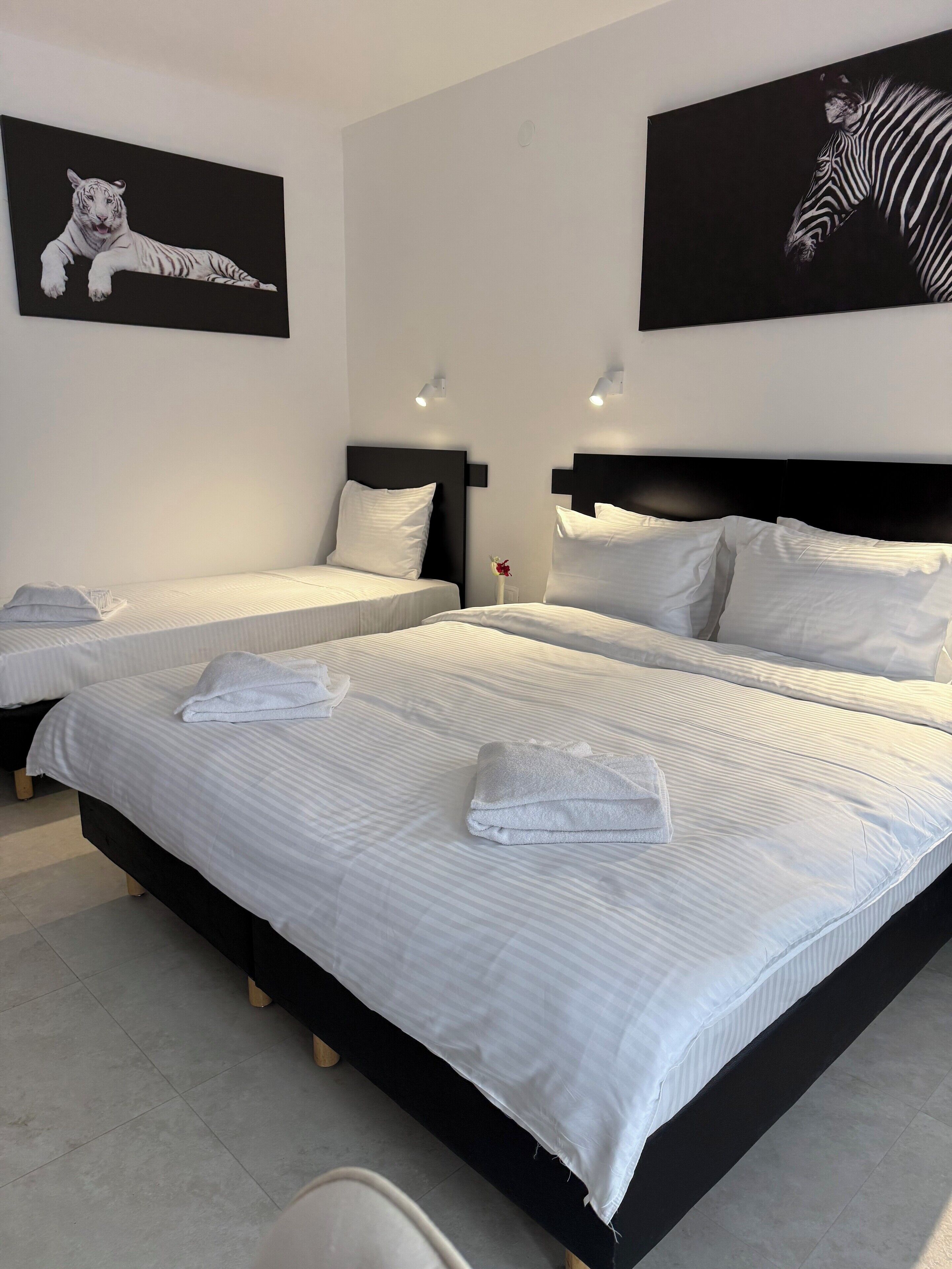 Quarto de hotel com duas camas, cada uma com lençóis e almofadas brancos, uma cama grande com uma toalha dobrada e duas pinturas emolduradas de animais nas paredes.