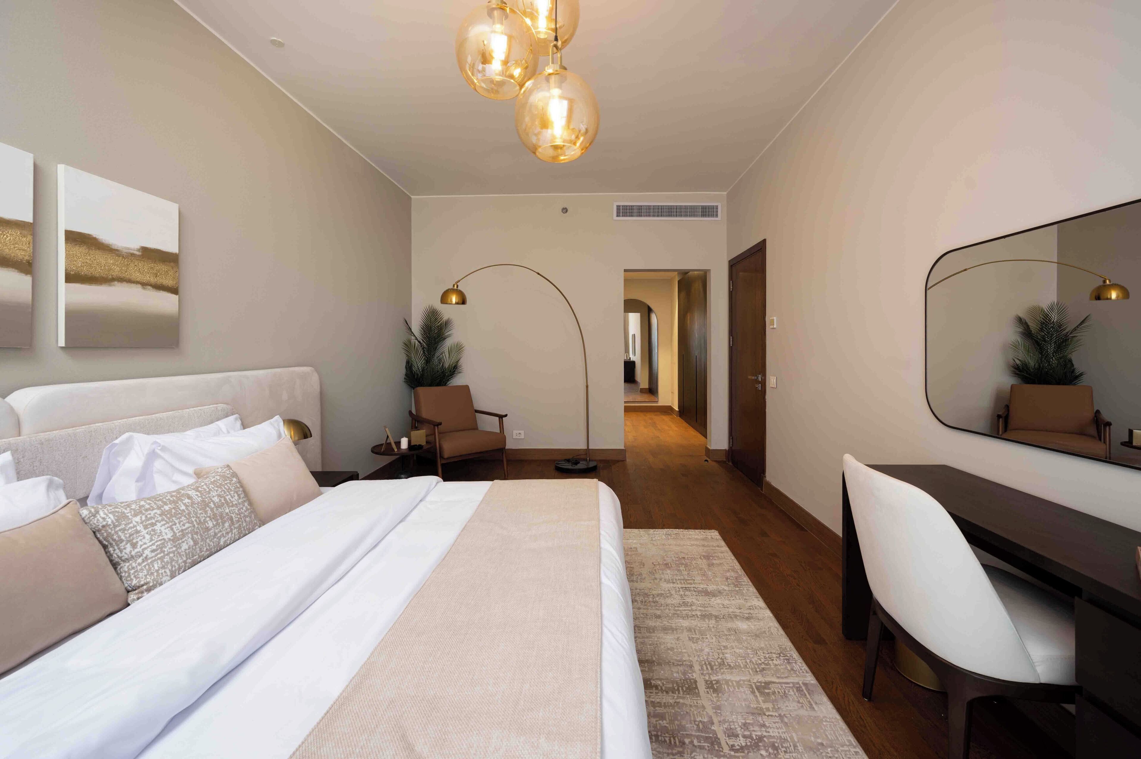 Quarto moderno com uma cama grande, uma escrivaninha e uma cadeira. Há um espelho na parede e um lustre no teto.