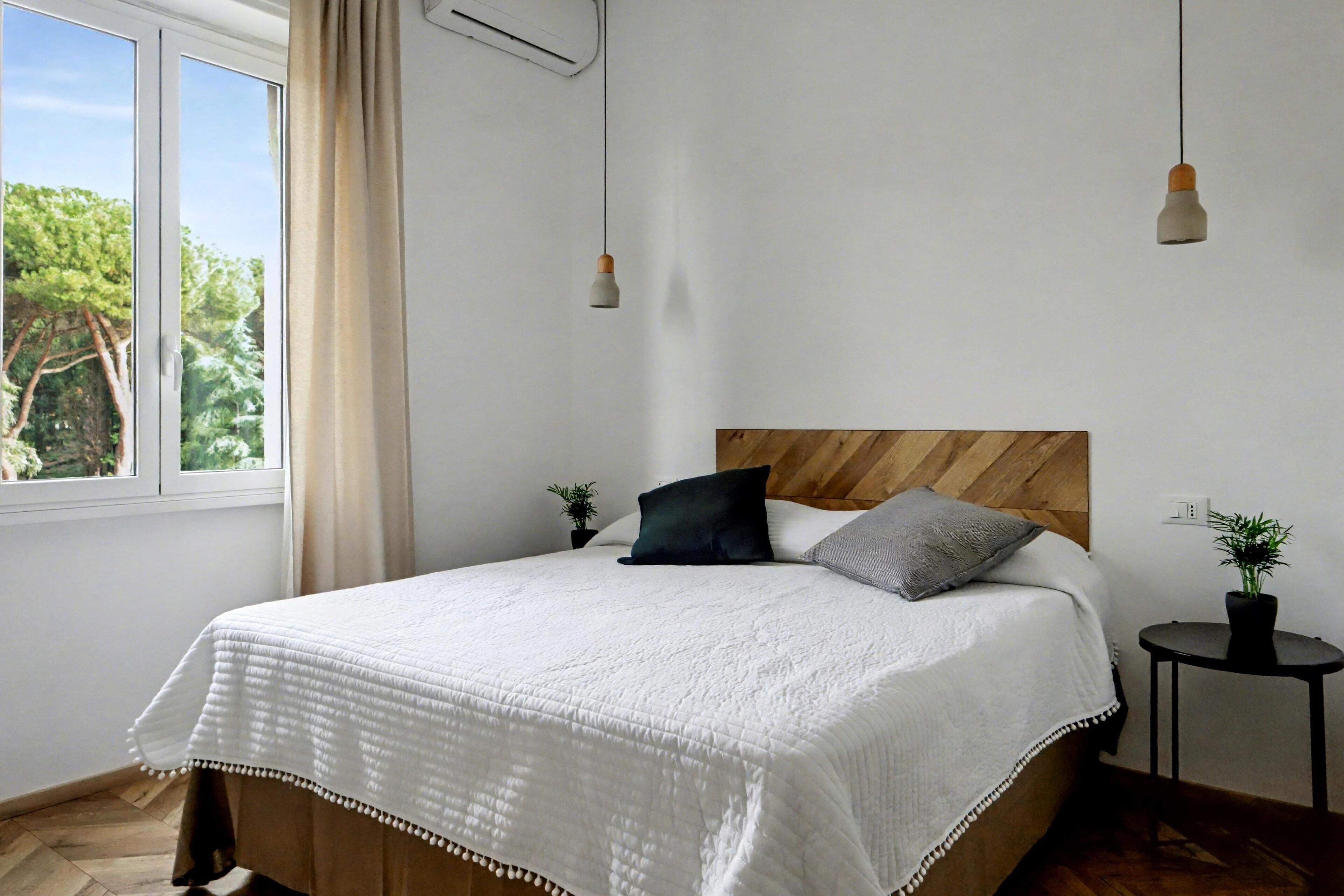 Quarto com cama, janela, planta em vaso e uma mesinha.