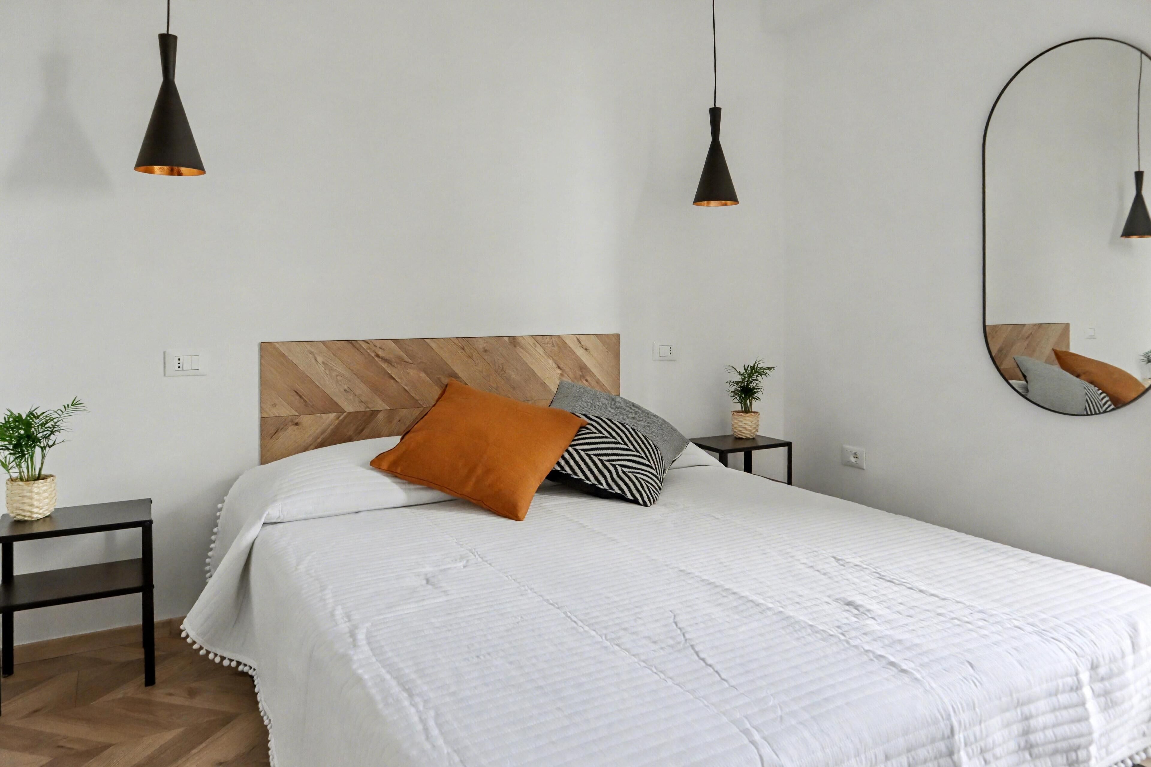 Um quarto moderno com cabeceira de madeira, roupa de cama branca e duas luminárias pendentes pretas.