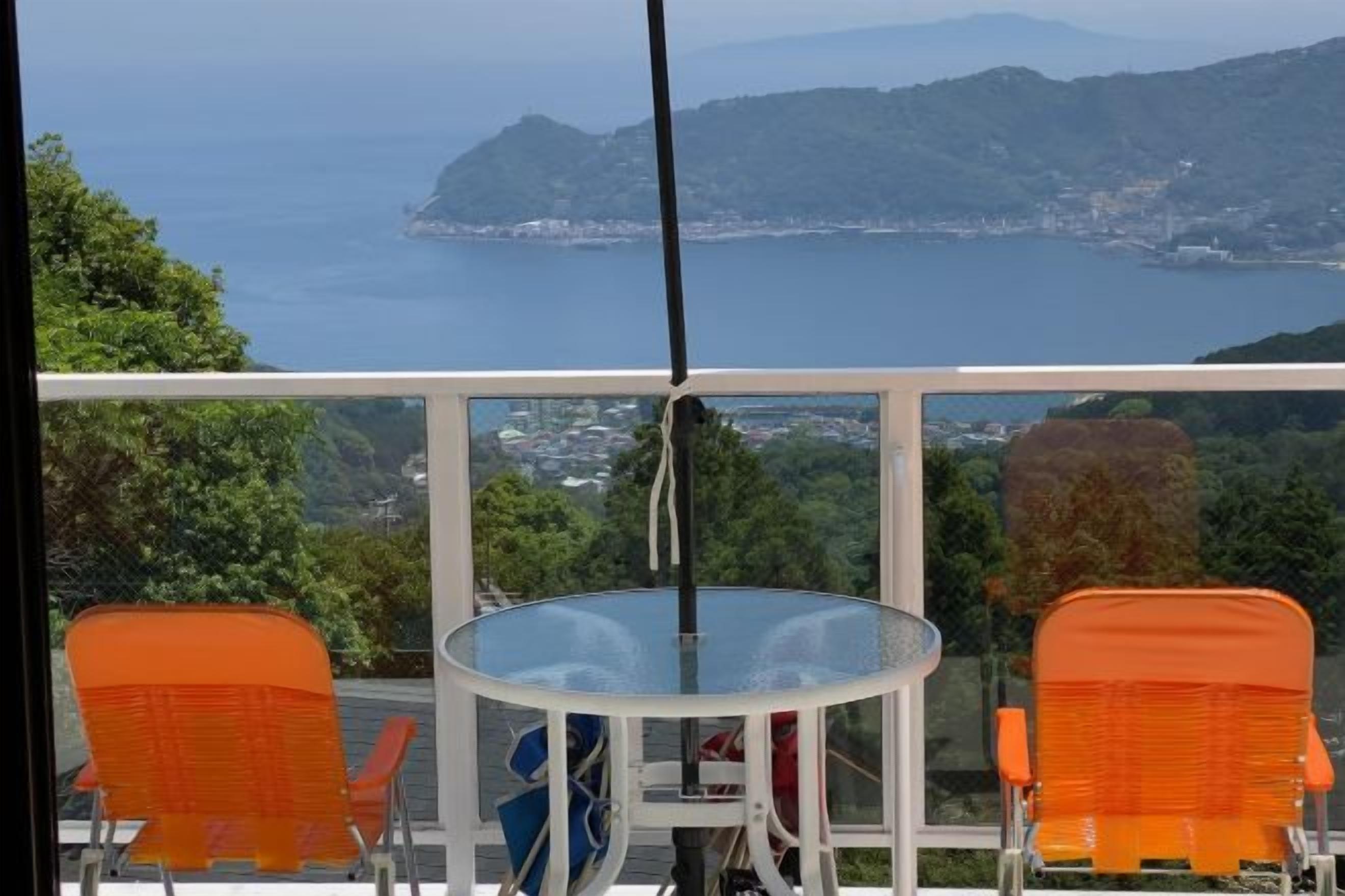 Varanda com mesa redonda e duas cadeiras laranja, com vista para paisagem costeira com colinas e um corpo de água.