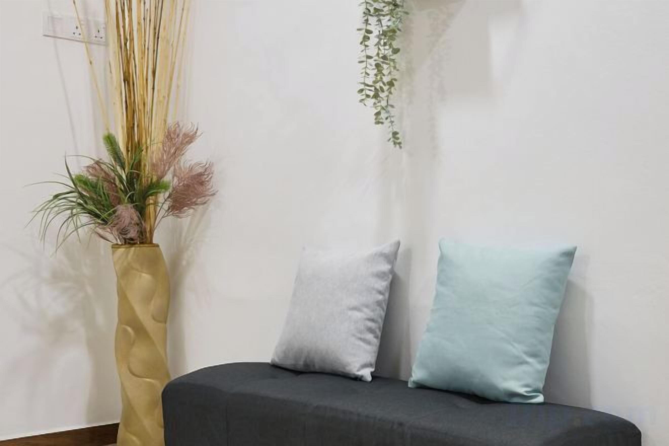 Quarto com sofá escuro em frente a uma parede branca, um vaso alto com plantas secas e uma planta pendente.