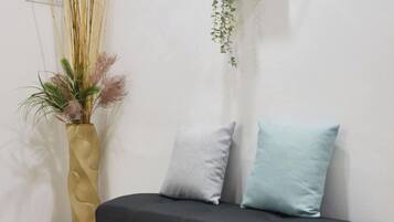 Quarto com sofá escuro em frente a uma parede branca, um vaso alto com plantas secas e uma planta pendente.