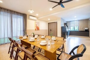 Uma sala de jantar com mesa de madeira posta para seis pessoas, rodeada por cadeiras de vime, em ambiente moderno com ventilador de teto e janelas amplas.