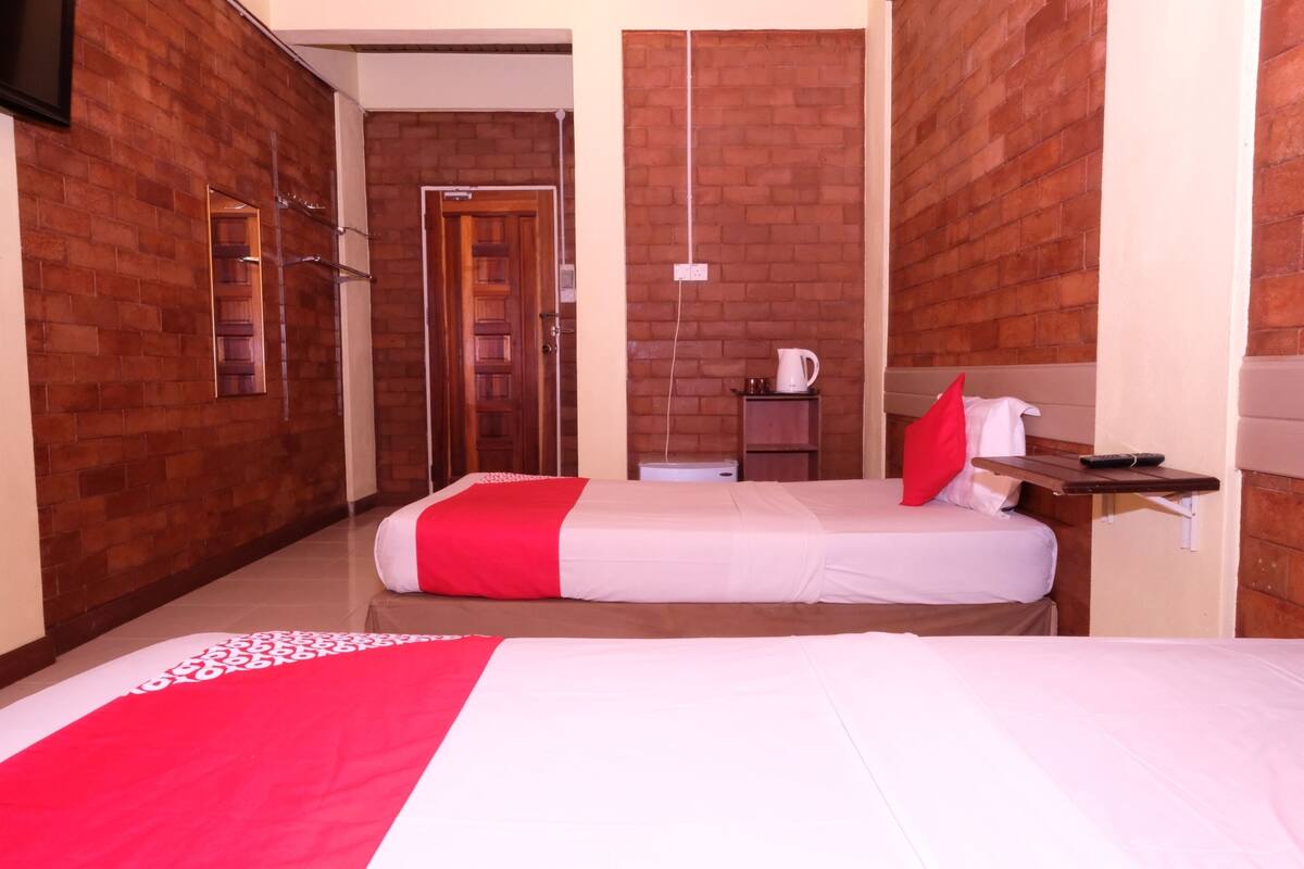 Quarto de hotel com duas camas, piso de madeira, parede de tijolos, uma mesinha com chaleira e um espelho na parede.