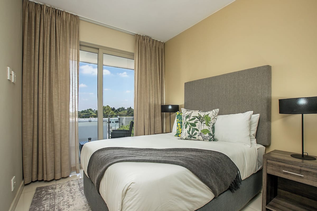Quarto de hotel com uma cama grande, uma mesa de cabeceira, um abajur e vista para o exterior.