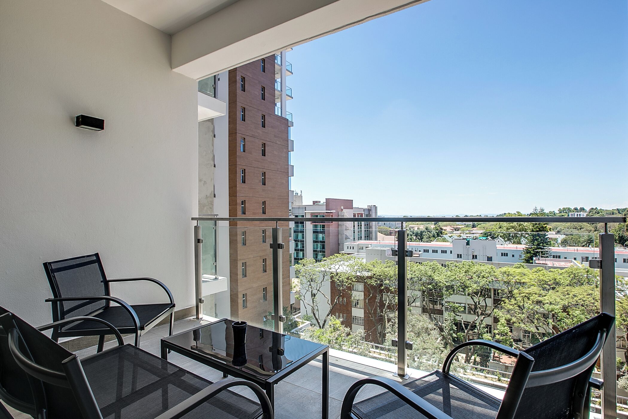 Uma varanda com mesa e cadeiras de vidro, oferecendo vista para uma paisagem urbana com edifícios e árvores.