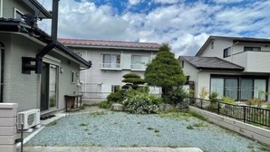 Zona residencial com uma casa moderna, varanda, pequeno jardim com pedras e plantas, e céu limpo.