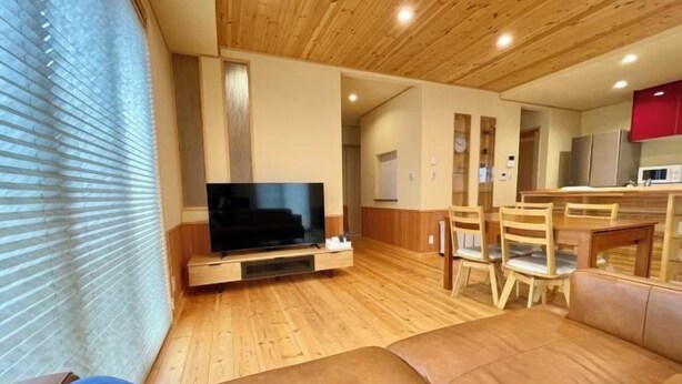 Uma sala de estar com televisão, piso de madeira, sofá e um espaço de refeições com cadeiras.