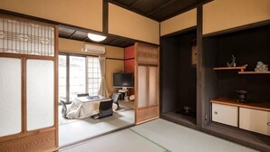 Um quarto japonês tradicional com tatames, uma escrivaninha com cadeira, uma televisão e uma estante com objetos decorativos.