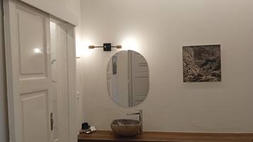 Quarto com cama, cómoda de madeira, espelho redondo, pia pequena e luminária fixada na parede.