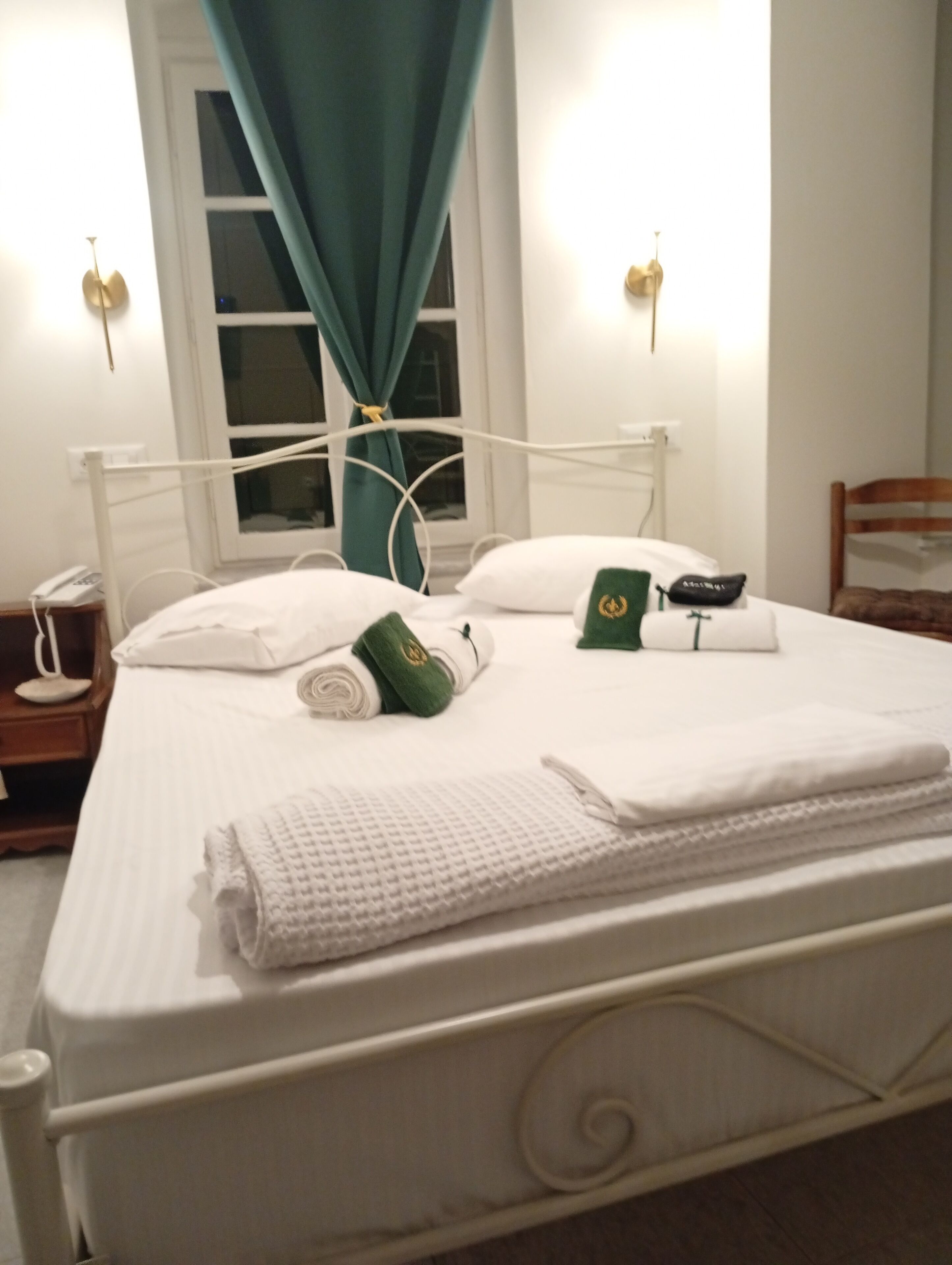 Cama bem arrumada com lençóis brancos e toalhas verdes, uma mesa de cabeceira com abajur, e uma janela com cortinas azul-turquesa.