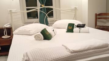 Cama bem arrumada com lençóis brancos e toalhas verdes, uma mesa de cabeceira com abajur, e uma janela com cortinas azul-turquesa.