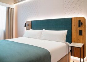 Quarto de hotel moderno com uma cama grande, cabeceira de madeira, mesa de cabeceira e luminária fixada na parede.