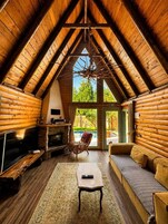 Interior de uma cabana de tronco com lareira, televisão, sofá e vista para o exterior.