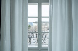 Vista através de uma janela com cortinas brancas, mostrando um edifício com vários andares e chão coberto de neve.