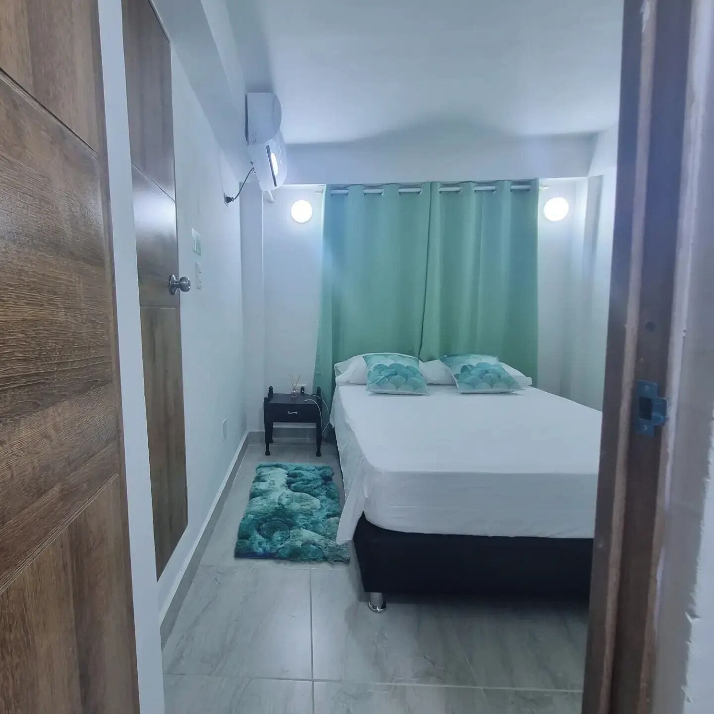 Quarto pequeno e bem arrumado, com uma cama, mesa de cabeceira, abajur e cortina verde.