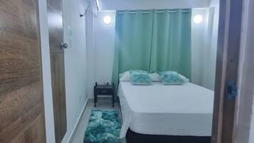 Quarto pequeno e bem arrumado, com uma cama, mesa de cabeceira, abajur e cortina verde.