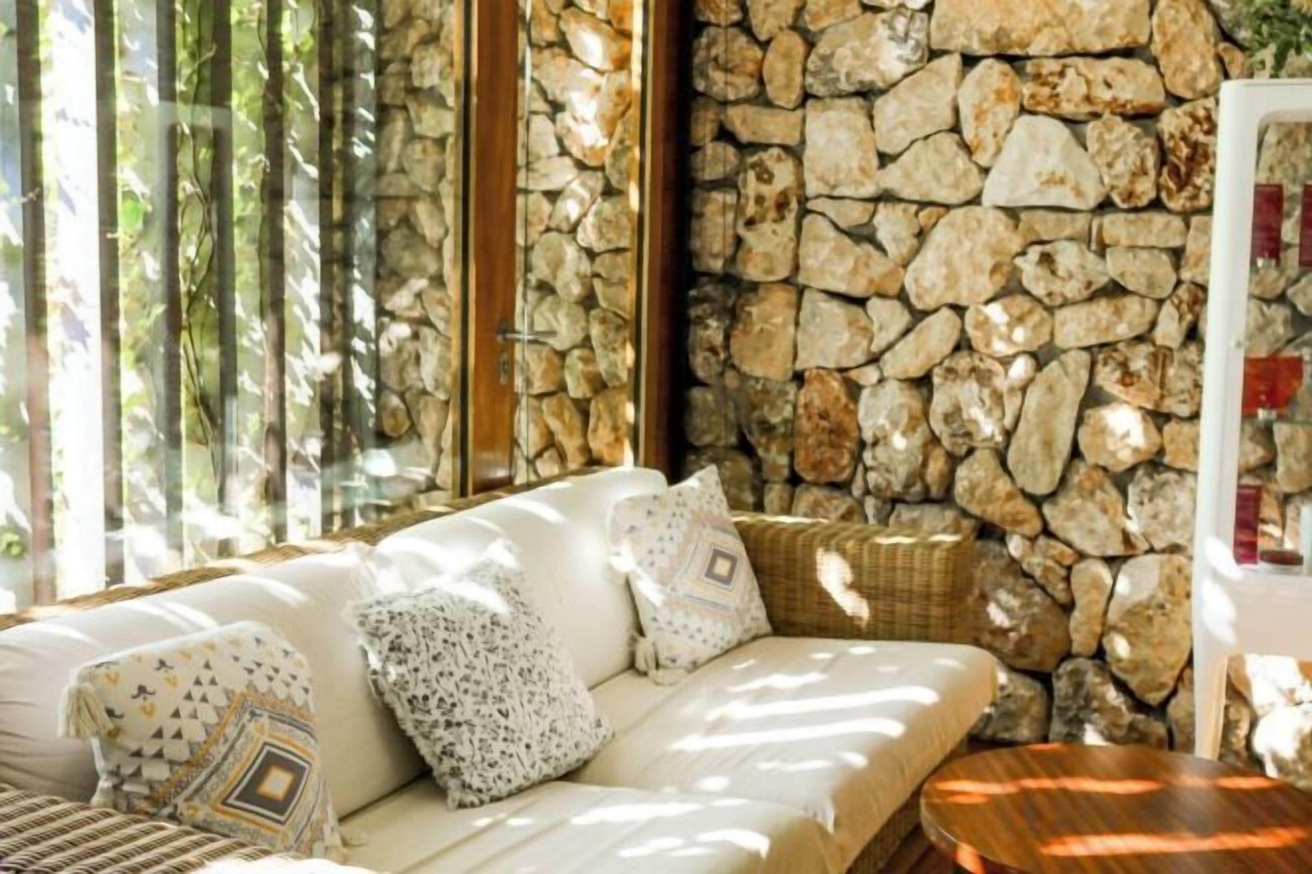 Um quarto com parede de pedra, um sofá branco com almofadas estampadas e uma cadeira de vime.