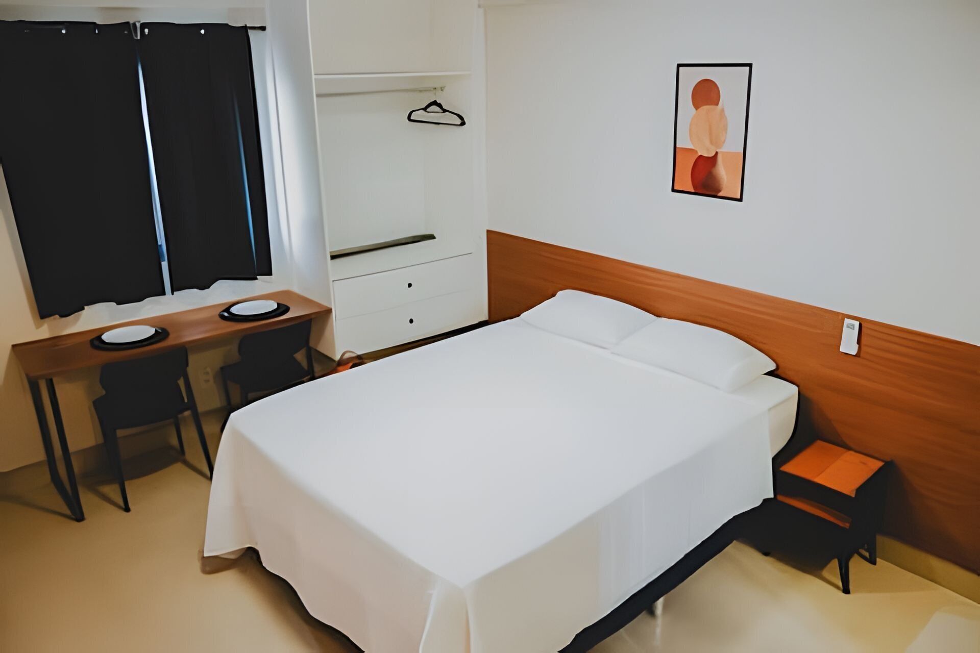 Quarto de hotel com cama, escrivaninha, cadeira, guarda-roupa e um quadro na parede.