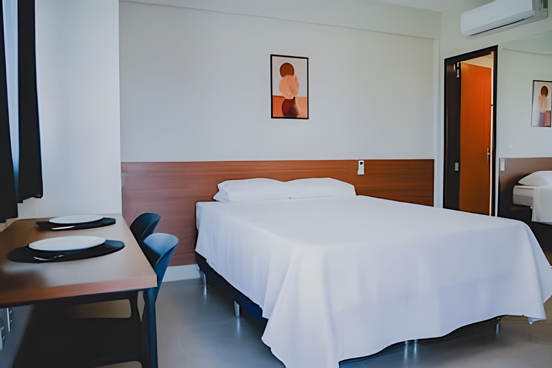 Quarto de hotel com uma cama, uma escrivaninha, uma cadeira e um quadro na parede.