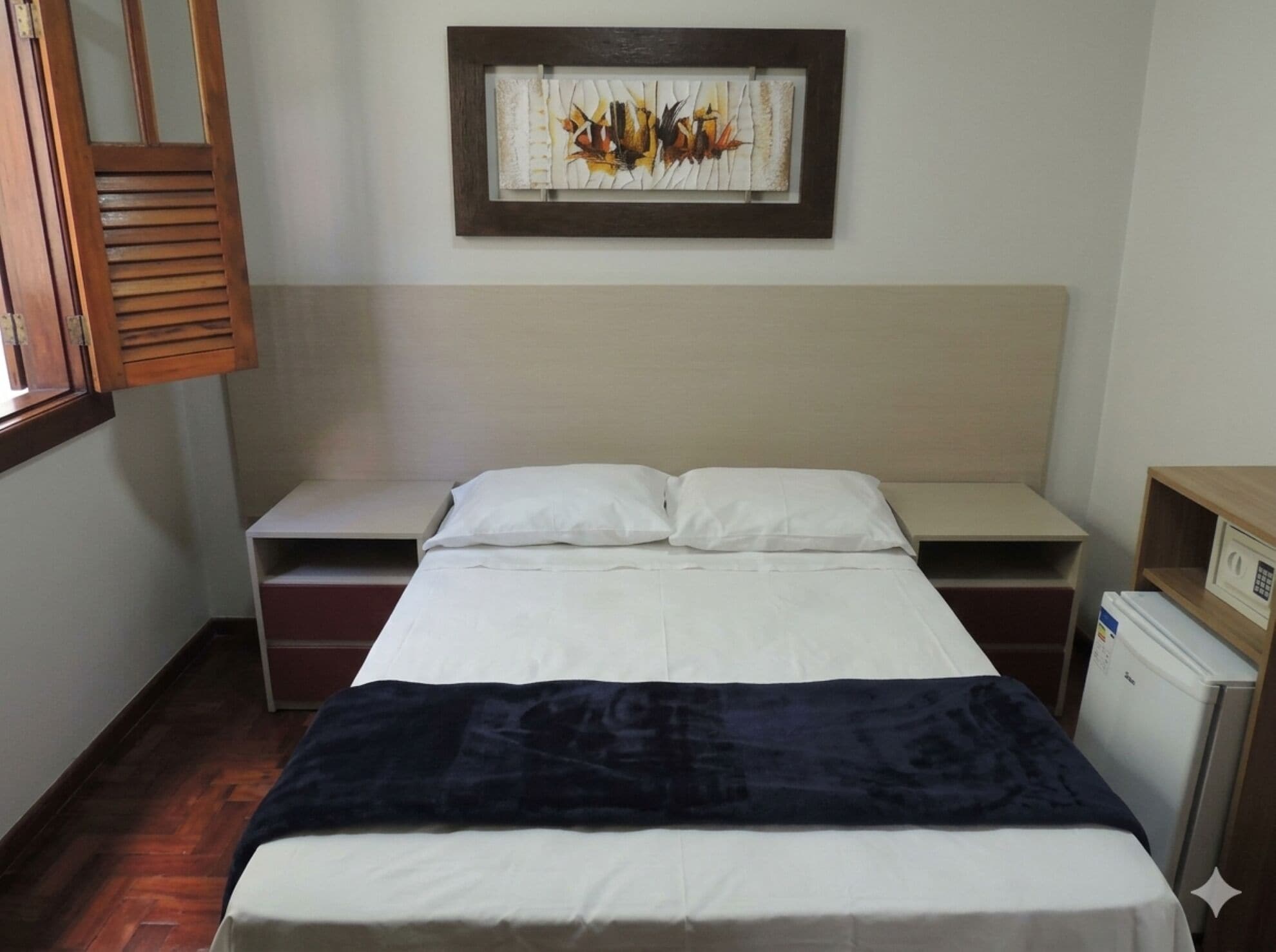 Quarto com uma cama, duas mesas de cabeceira, um criado-mudo, um armário de madeira e uma obra de arte na parede.
