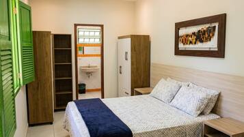 Quarto com cama, guarda-roupa, mesa de cabeceira, quadro na parede e banheiro visível através de uma porta aberta.
