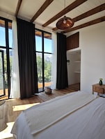 Quarto com uma cama grande, vigas de madeira, luminária pendente, vista para montanhas e piso de madeira.