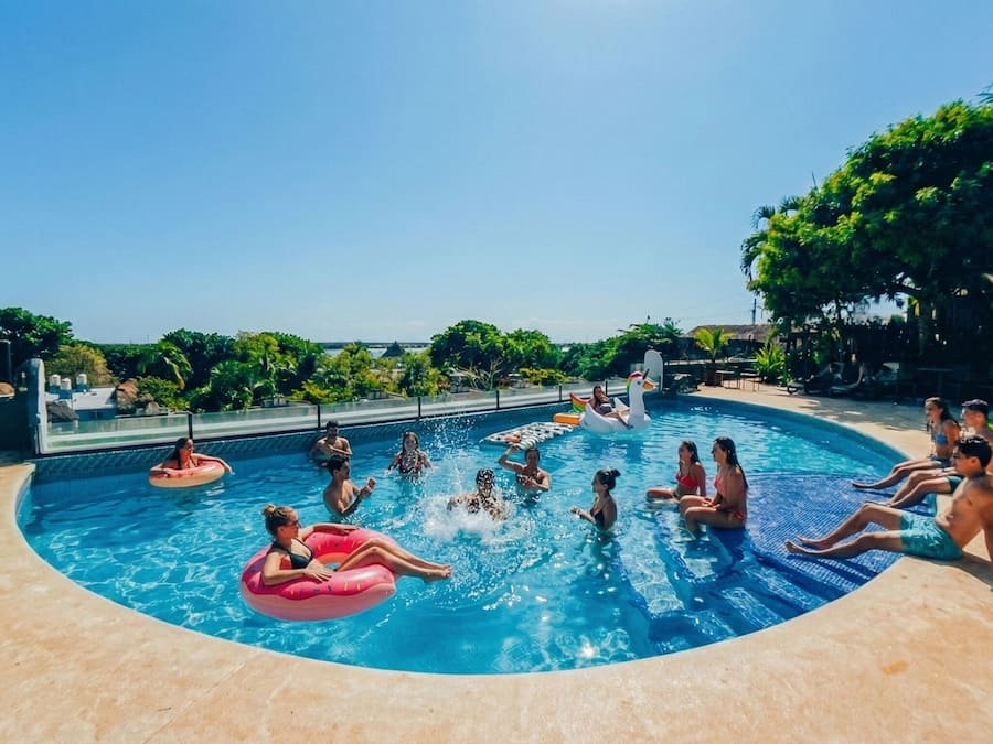 Uma área de piscina com pessoas nadando e relaxando, cercada por palmeiras e um céu claro.