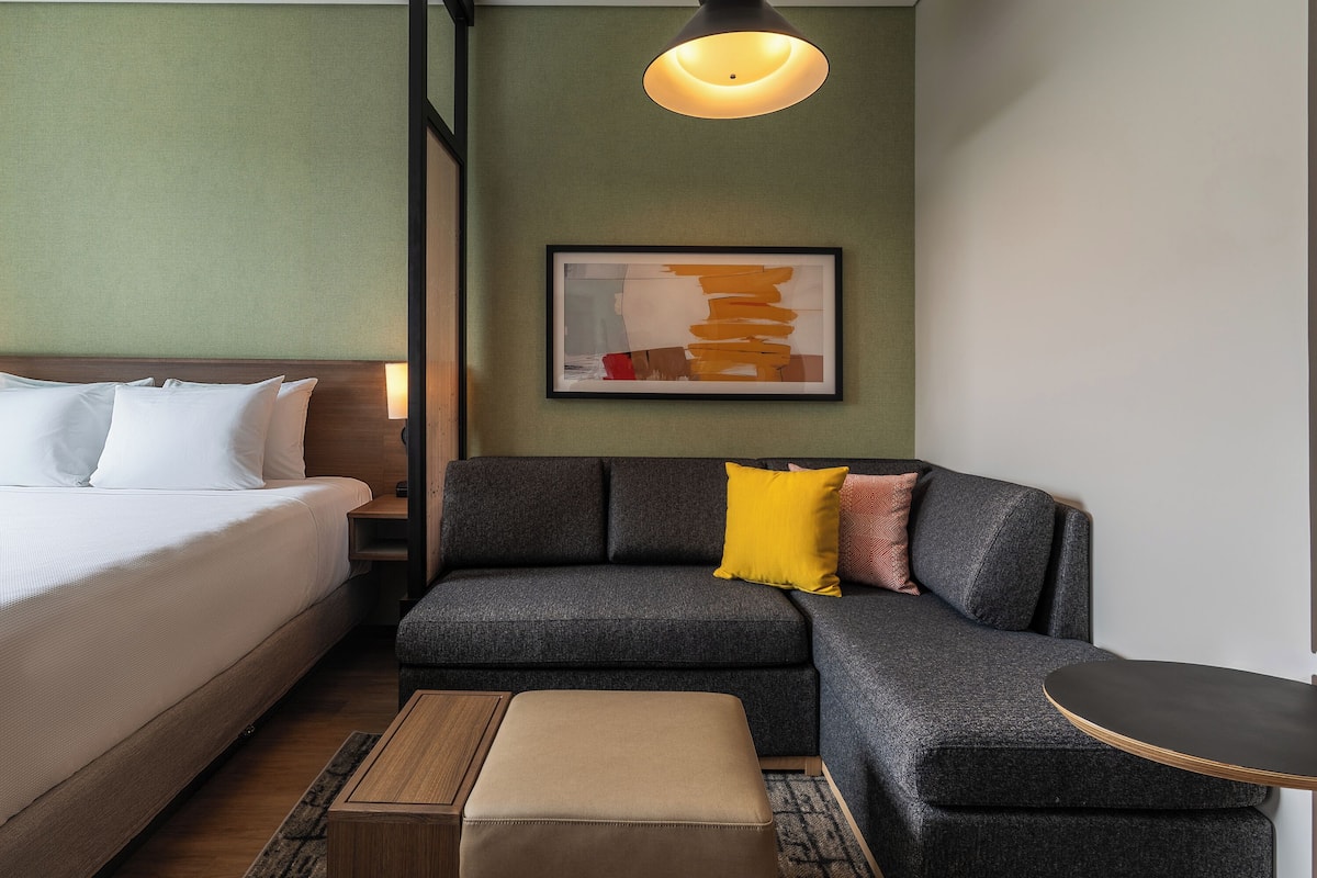 Quarto de hotel com uma cama, um sofá cinzento com almofadas amarelas e vermelhas, uma mesinha redonda e uma pintura abstrata emoldurada na parede.