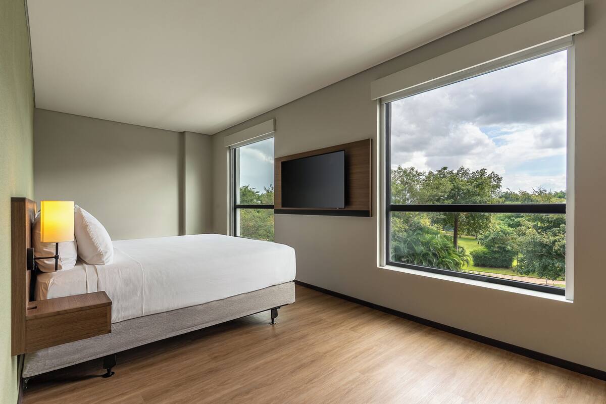 Quarto moderno com uma janela grande, uma cama, uma mesa de cabeceira e uma TV fixada na parede.