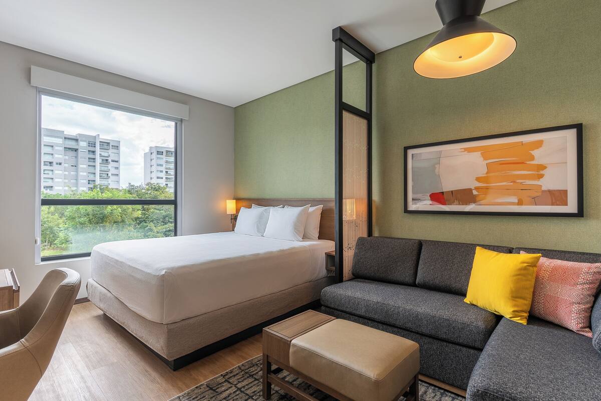 Quarto de hotel moderno com uma cama grande, um sofá, uma mesa pequena e vista para prédios pela janela.