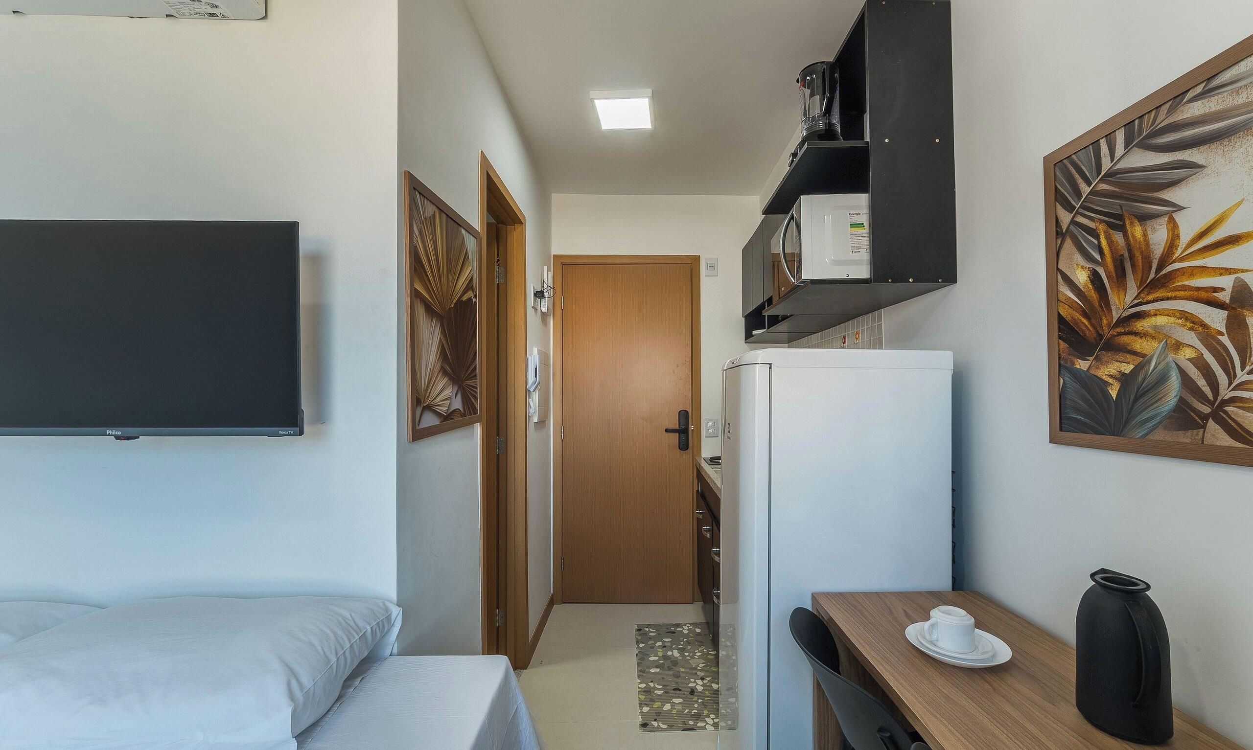 Quarto de hotel pequeno com uma cama, uma mesinha com uma xícara e um vaso, um refrigerador, um micro-ondas e uma TV fixada na parede.