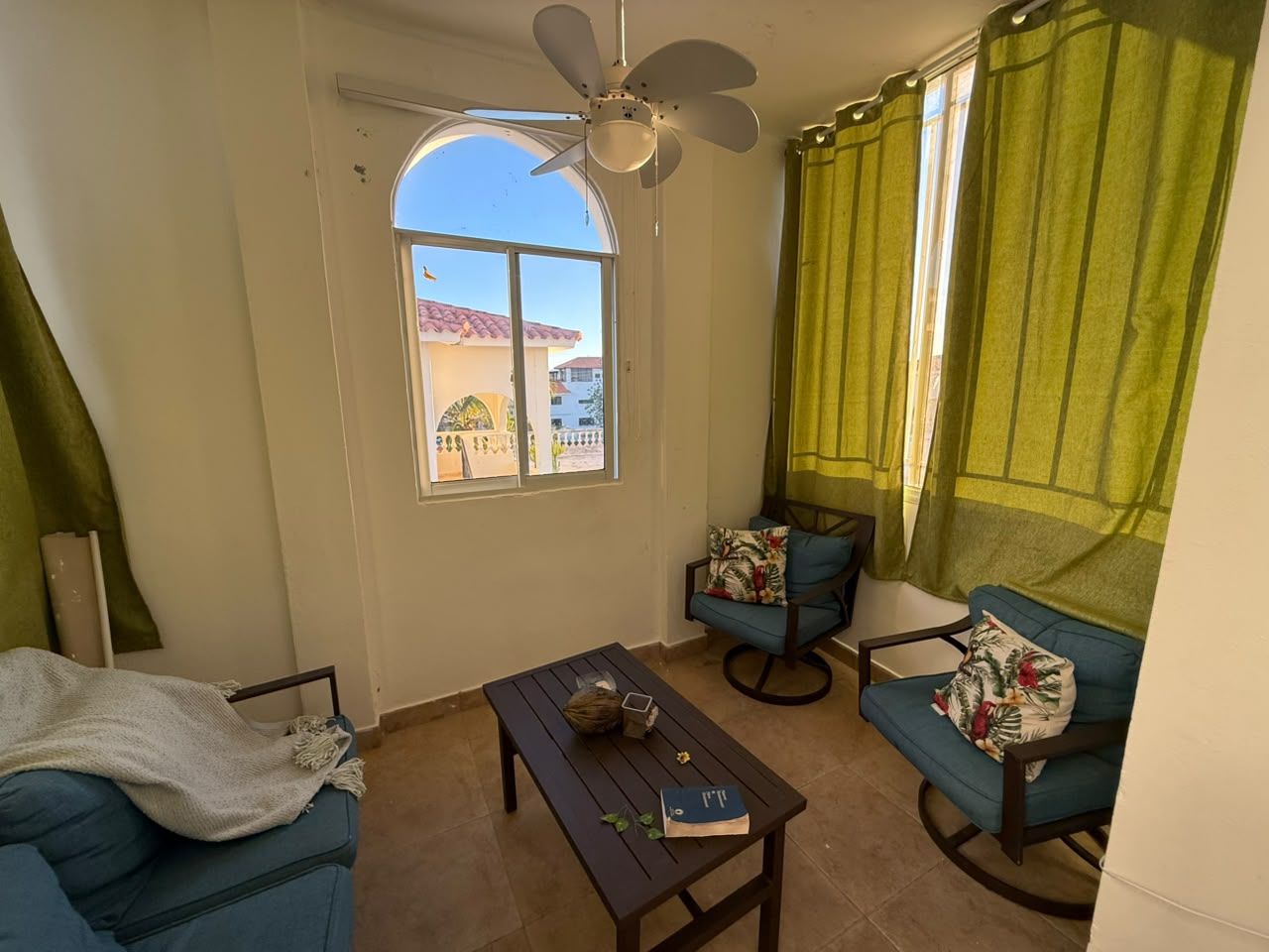 Quarto com ventoinha no teto, janela com vista, sofá, cadeira e mesa pequena.