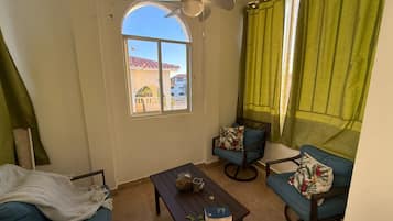 Quarto com ventoinha no teto, janela com vista, sofá, cadeira e mesa pequena.