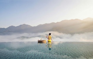 Pessoa de biquíni amarelo sentada em piscina infinita com água cristalina, cercada por montanhas e névoa.