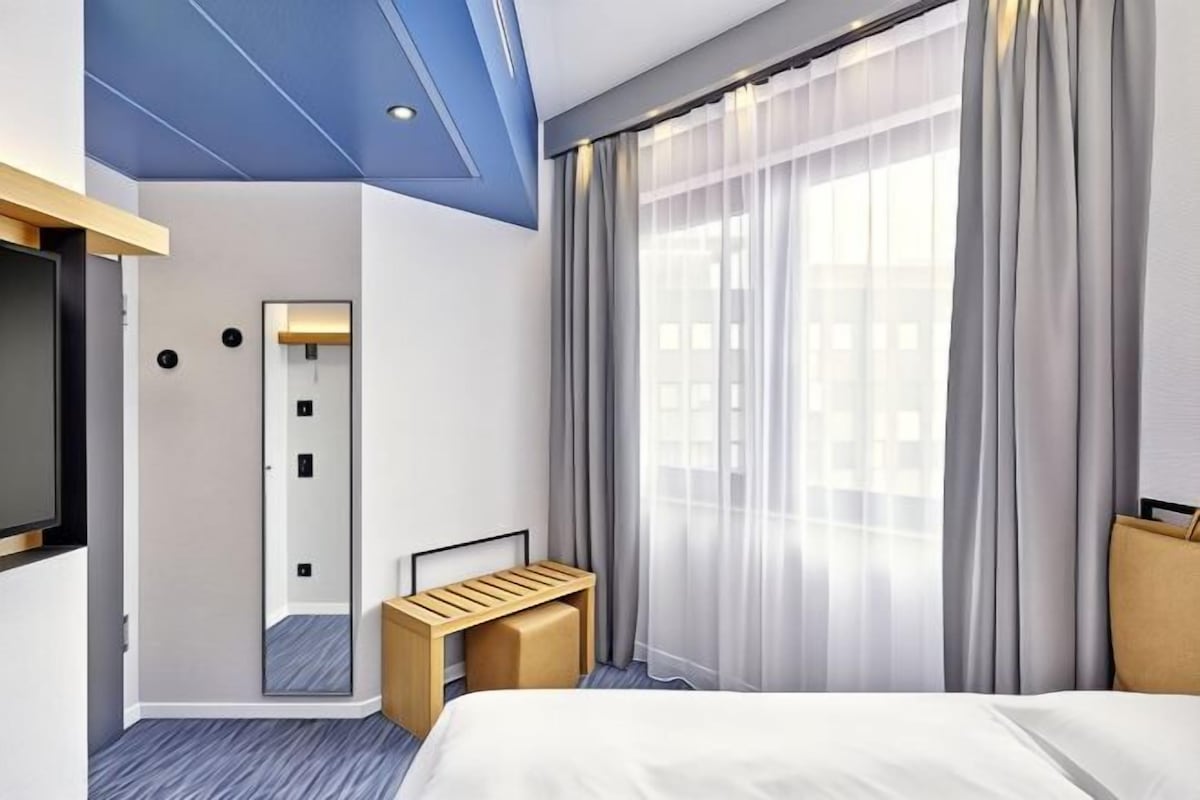 Quarto de hotel com uma cama, uma mesa, um espelho e uma janela com cortinas.