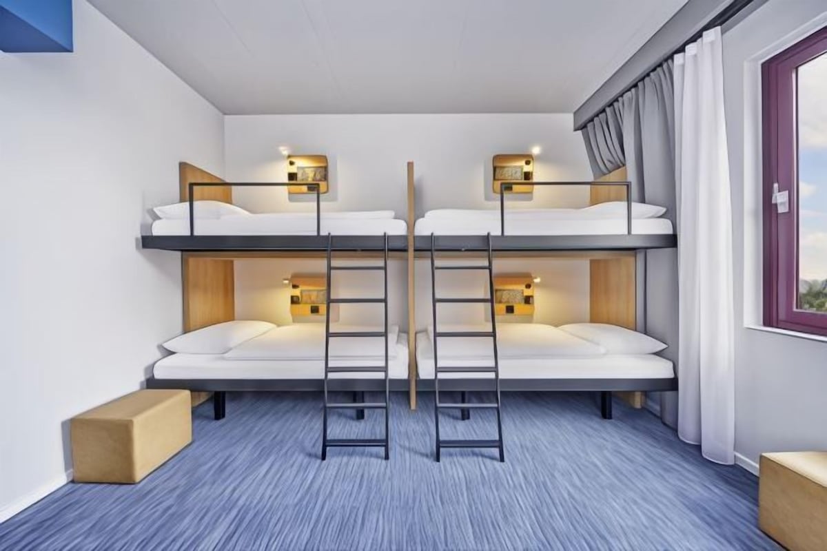 Quarto com beliches, duas mesas de cabeceira com luminárias, uma janela com cortinas e um tapete azul.